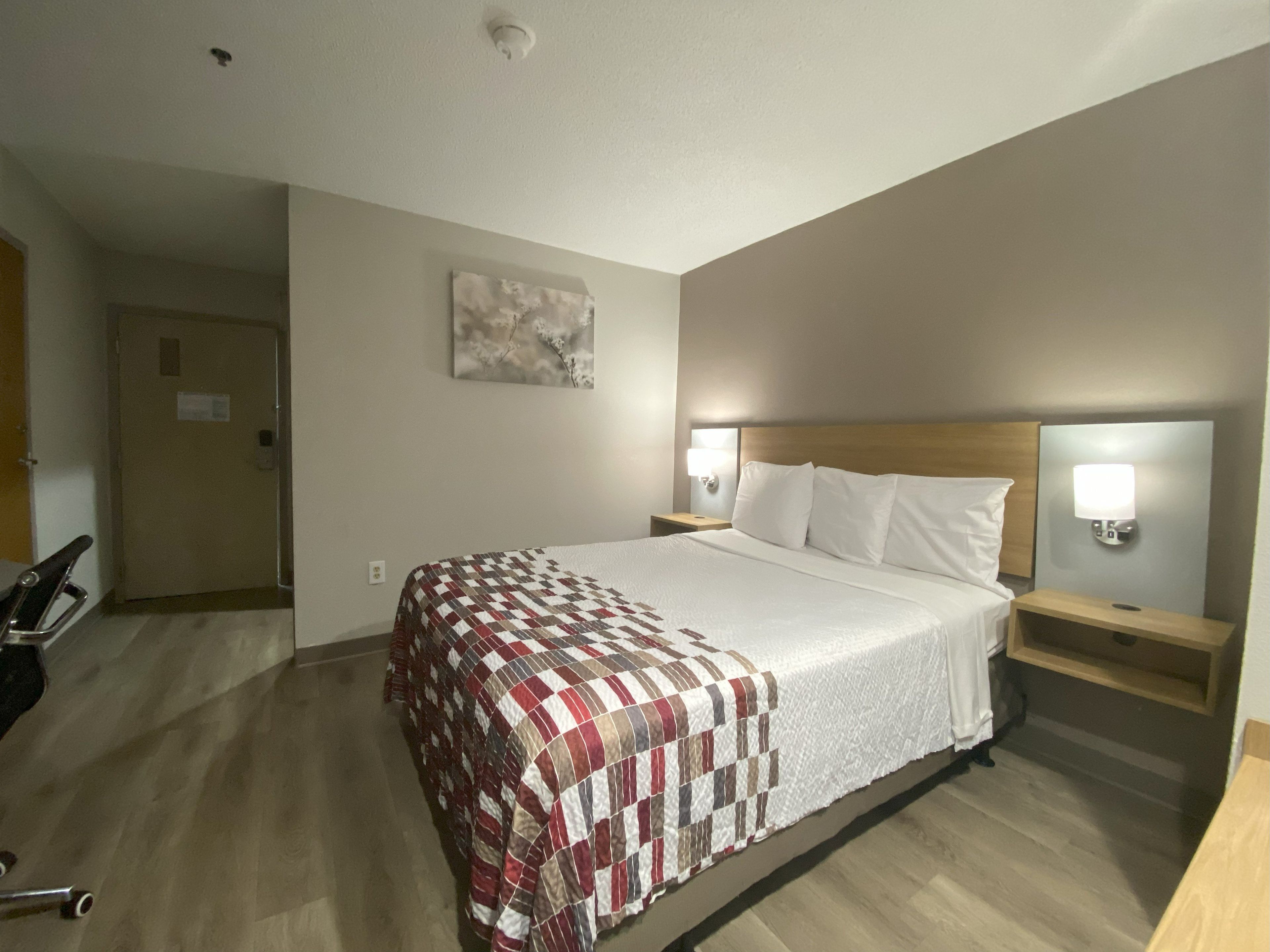 Quarto de hotel com uma cama grande, cabeceira de madeira, duas mesas de cabeceira com abajures, uma porta e uma obra de arte na parede.