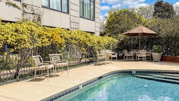 Uma área de piscina com piscina azul cristalina, rodeada por um terraço com cadeiras e mesas, e uma árvore florida amarela ao fundo.