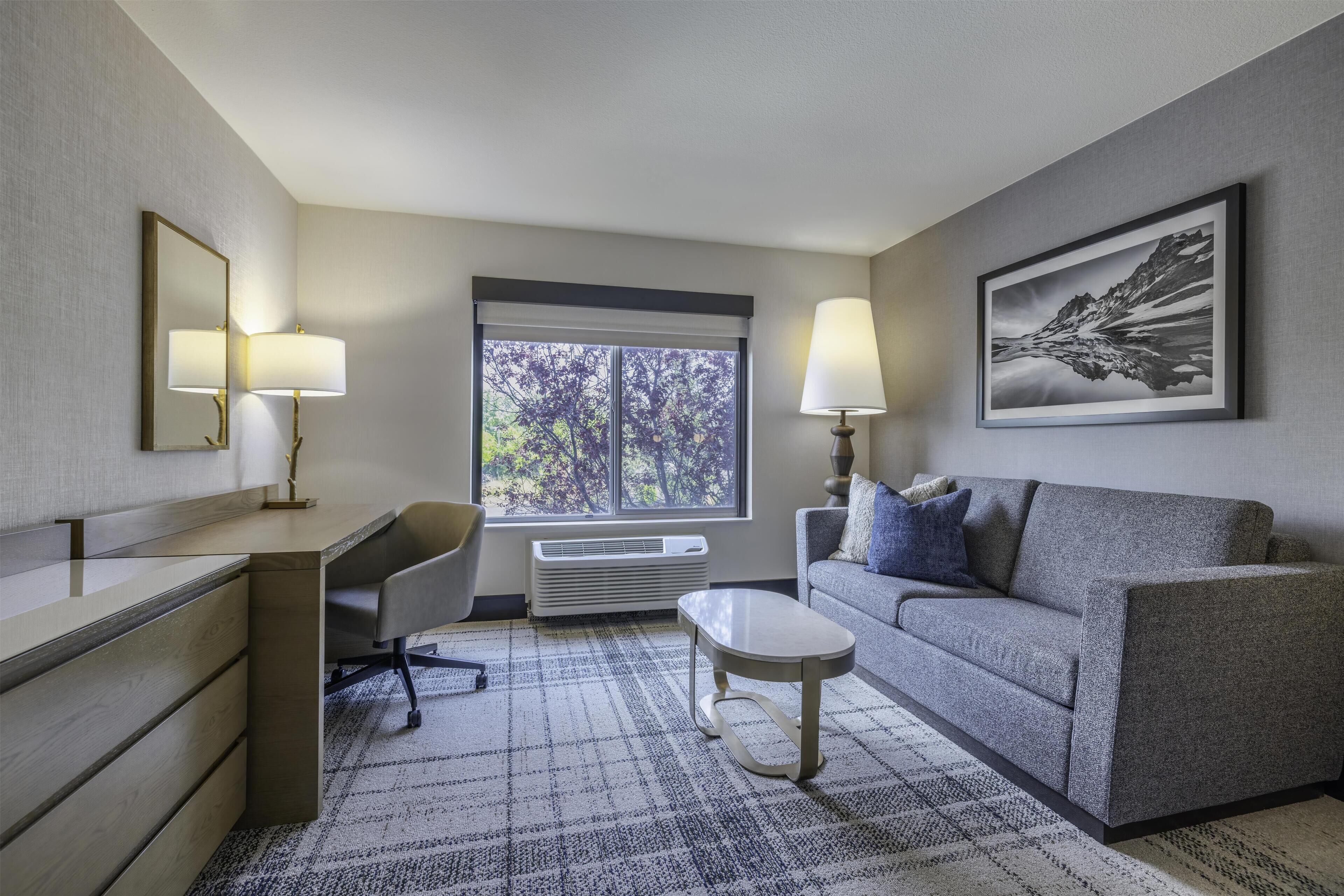 Quarto de hotel com escrivaninha, cadeira, sofá e uma grande janela com vista para árvores.