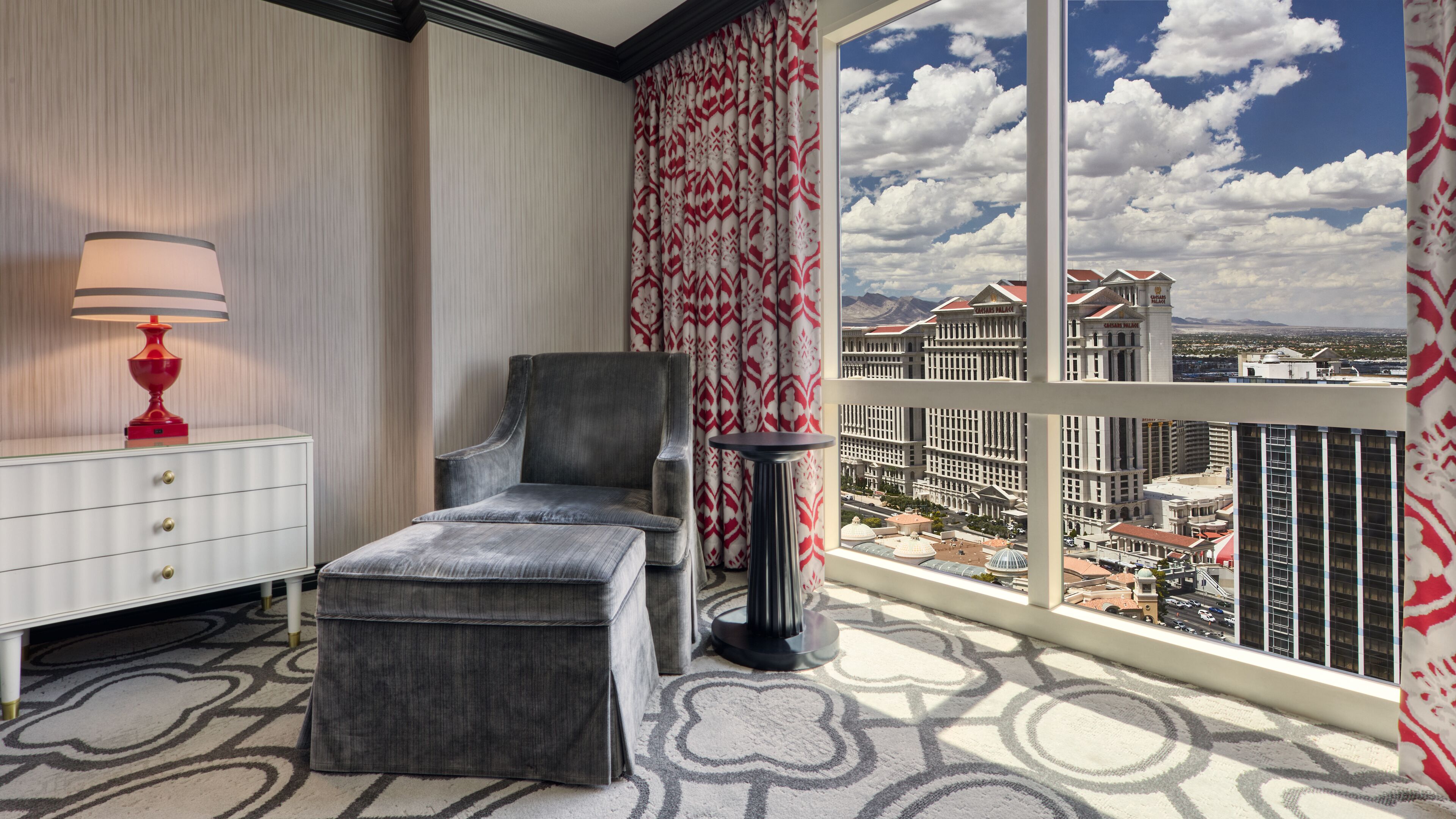 Quarto de hotel com uma janela grande, um abajur vermelho, uma poltrona cinza e um penteado branco.