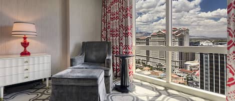 Quarto de hotel com uma janela grande, um abajur vermelho, uma poltrona cinza e um penteado branco.