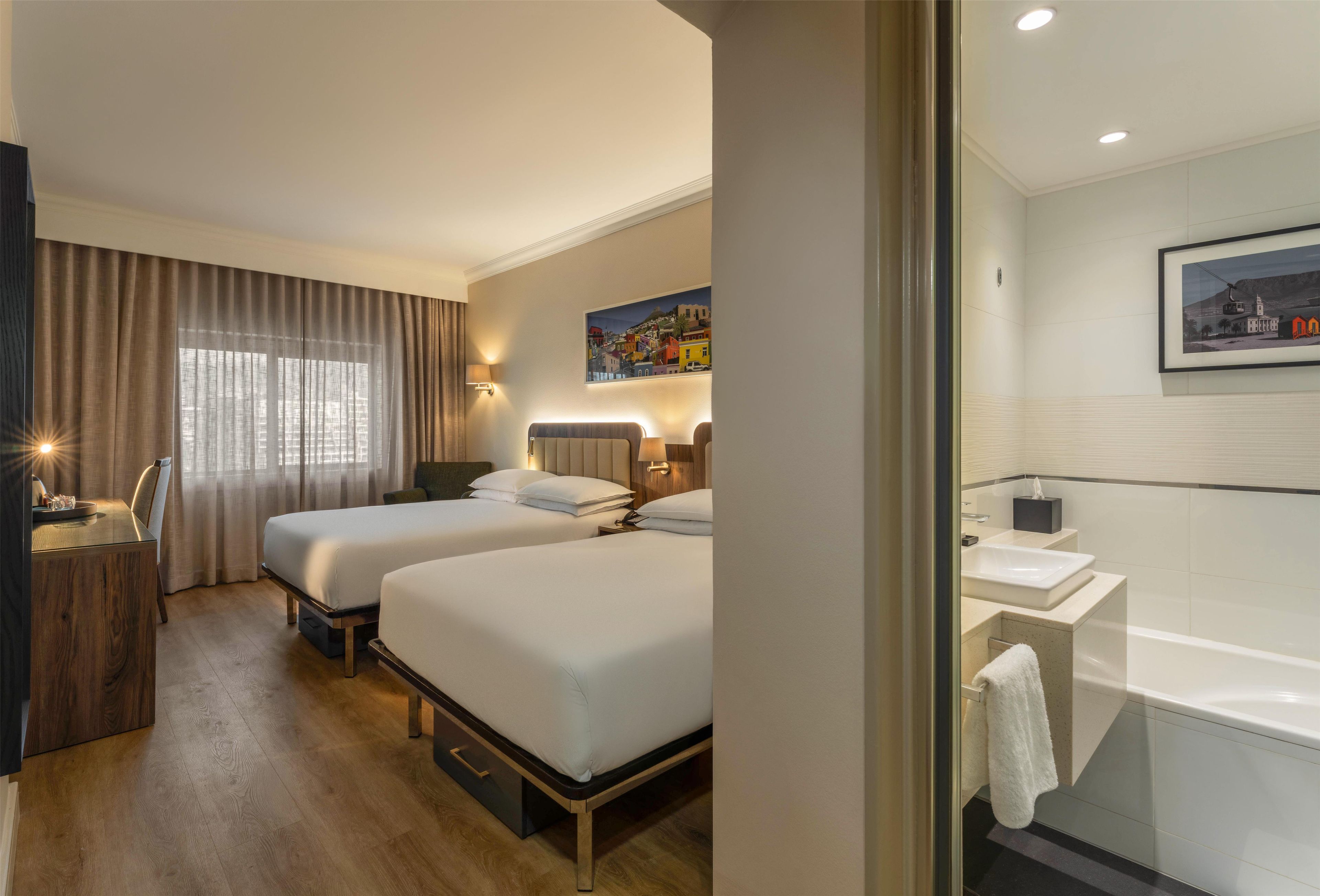Quarto de hotel com duas camas, uma escrivaninha, uma cadeira, janela com cortinas, um quadro na parede e banheiro com banheira e pia.