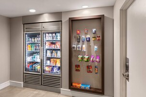 Quarto de hotel com minibar, expositor de snacks na parede e porta com puxador.