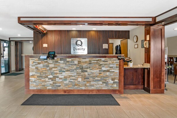 Uma mesa de recepção com a placa 'Quality by Choice Hotels', um quiosque de check-in digital e um espaço de atendimento em madeira.