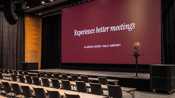 Uma sala de conferências com uma tela grande exibindo o texto 'Experience better meetings' e 'Clarion Hotel Oslo Airport'. Fileiras de cadeiras organizadas para os participantes.