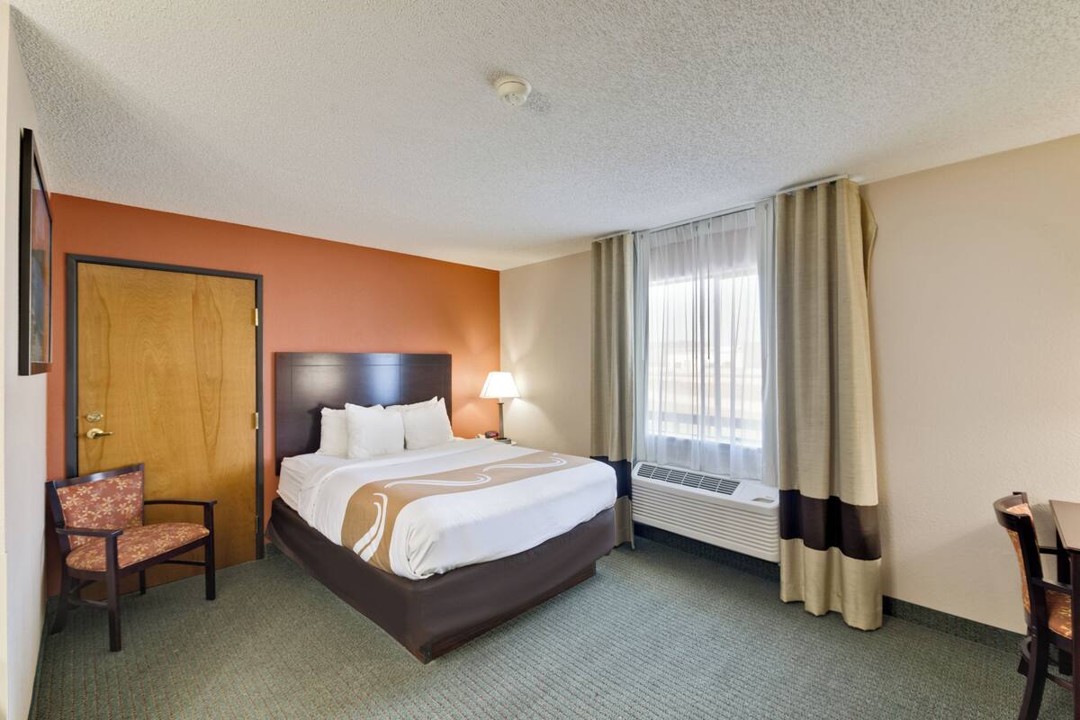 Quarto de hotel com uma cama grande, uma cadeira, uma escrivaninha e uma janela com cortinas.