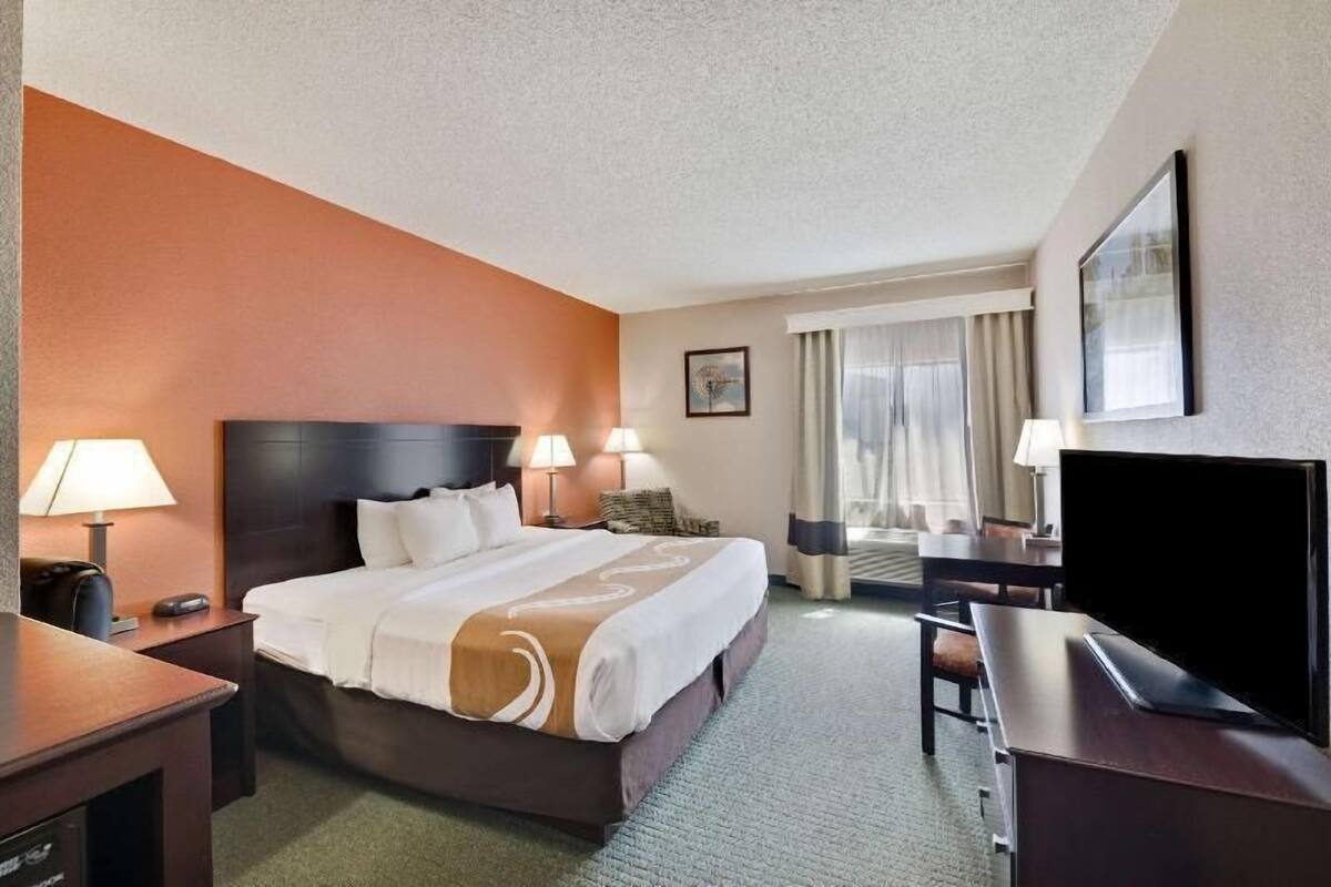 Quarto de hotel com cama, luminárias de cabeceira, escrivaninha, cadeira, televisão e janela com cortinas.