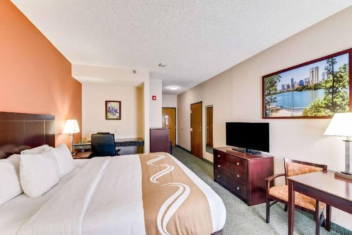 Quarto de hotel com cama, escrivaninha, cadeira, cômoda, televisão e um quadro na parede.
