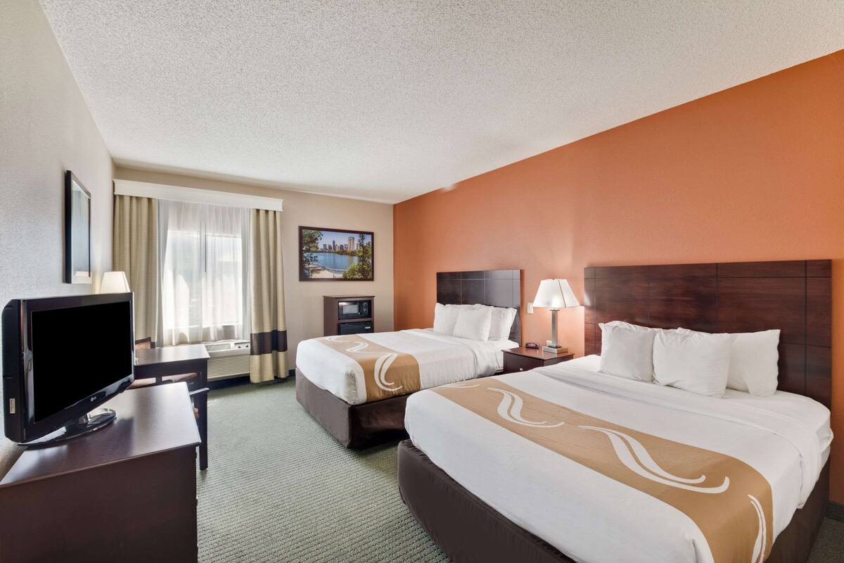 Quarto de hotel com duas camas, televisão, escrivaninha e janela com cortinas.