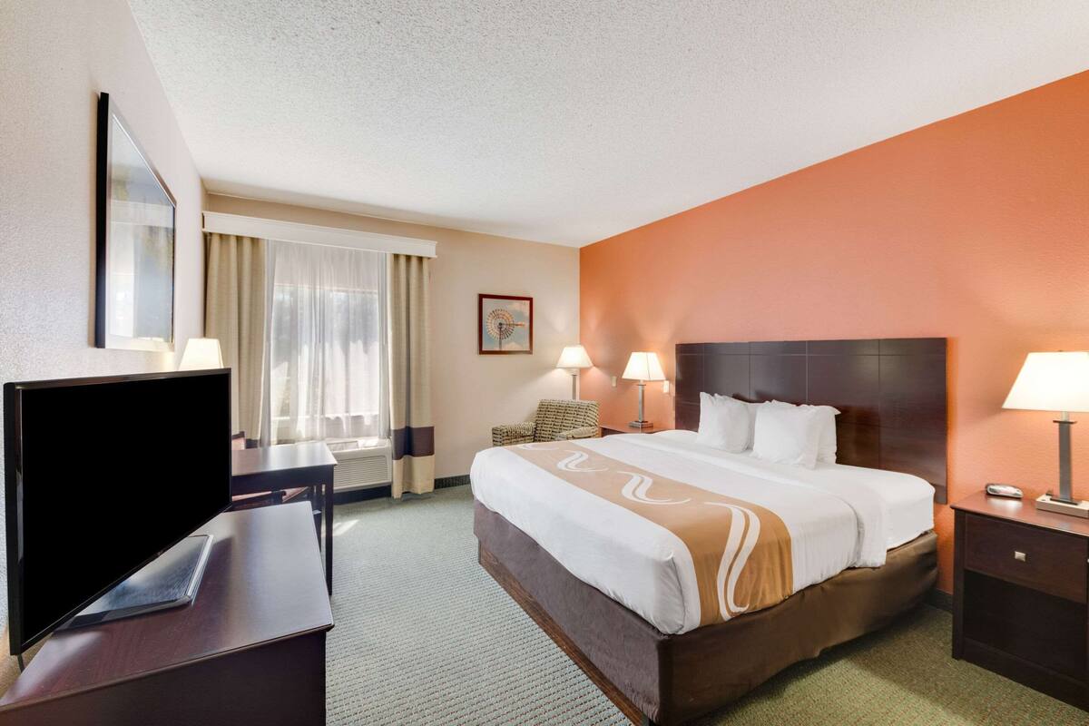 Quarto de hotel com uma cama grande, televisão de tela plana, escrivaninha com abajur e janela com cortinas.
