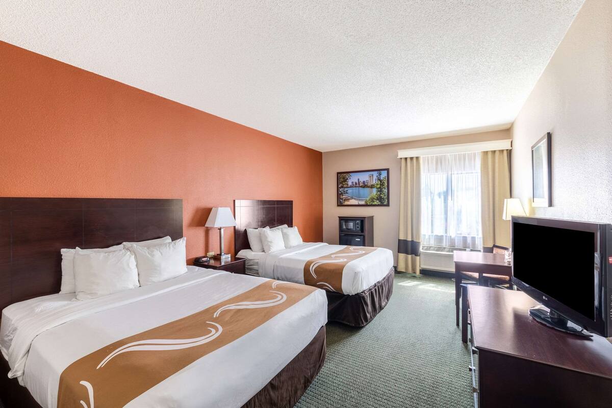 Quarto de hotel com duas camas, uma escrivaninha com televisão, um quadro na parede e uma janela com cortinas.