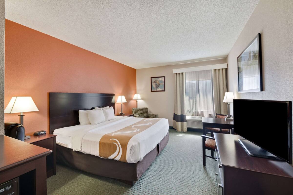 Quarto de hotel com uma cama grande, duas mesas de cabeceira com abajures, uma escrivaninha com televisão de tela plana e uma janela com cortinas.