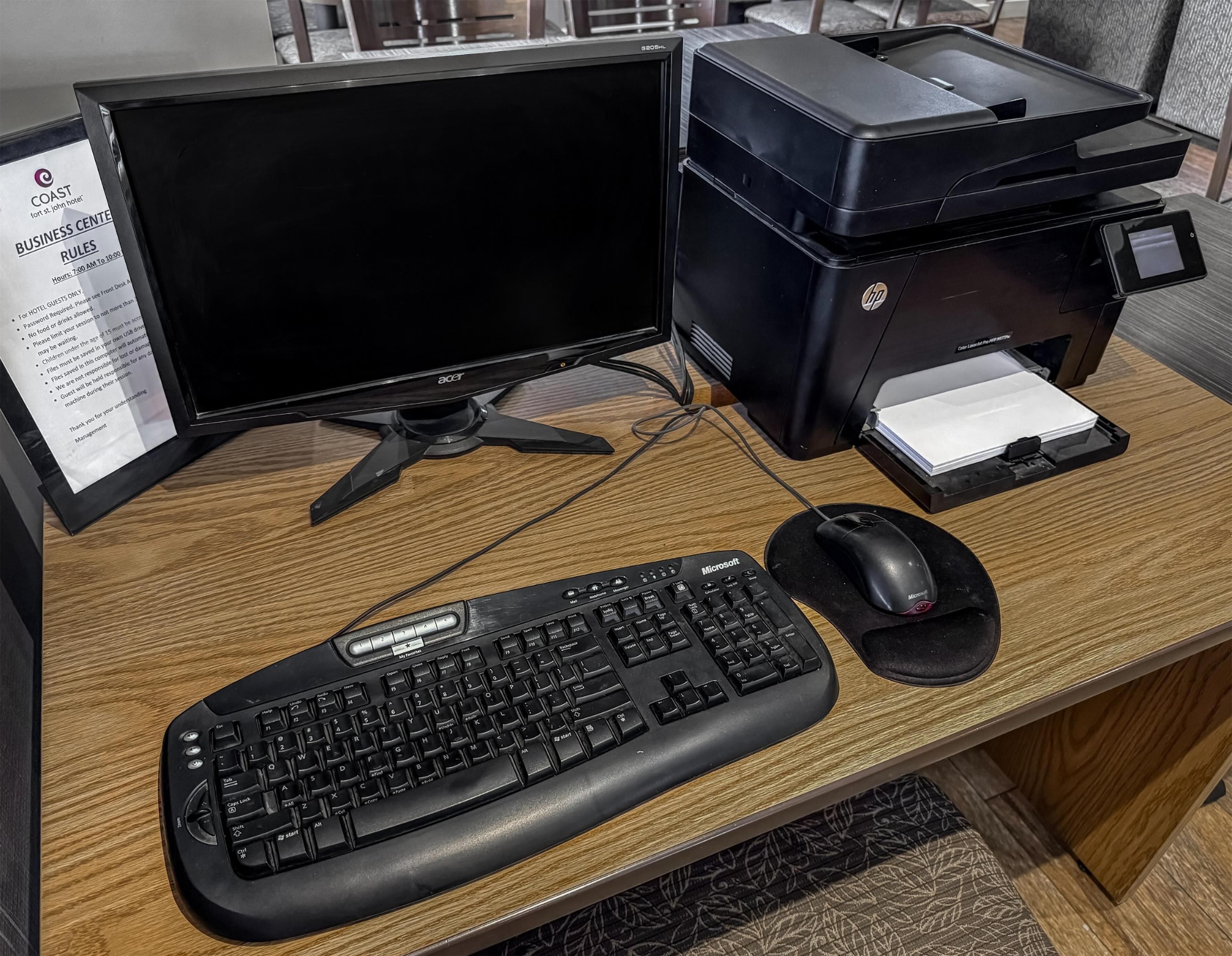 Mesa de centro de negócios em hotel, com monitor Acer, impressora HP, teclado Microsoft e rato.