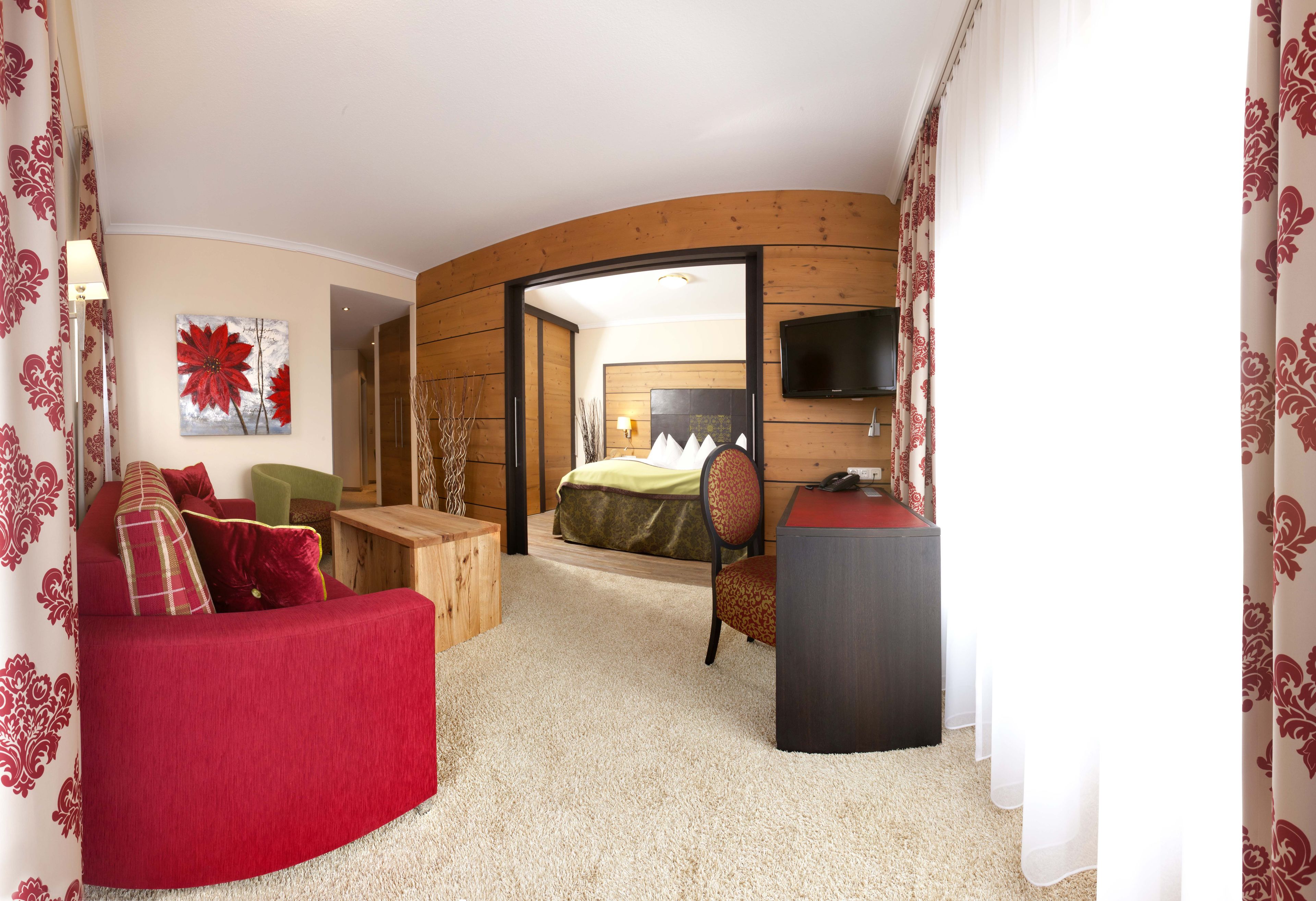 Quarto de hotel moderno com uma parede curva de madeira, uma cama com cabeceira verde, um sofá vermelho, uma escrivaninha e uma TV de tela plana na parede.