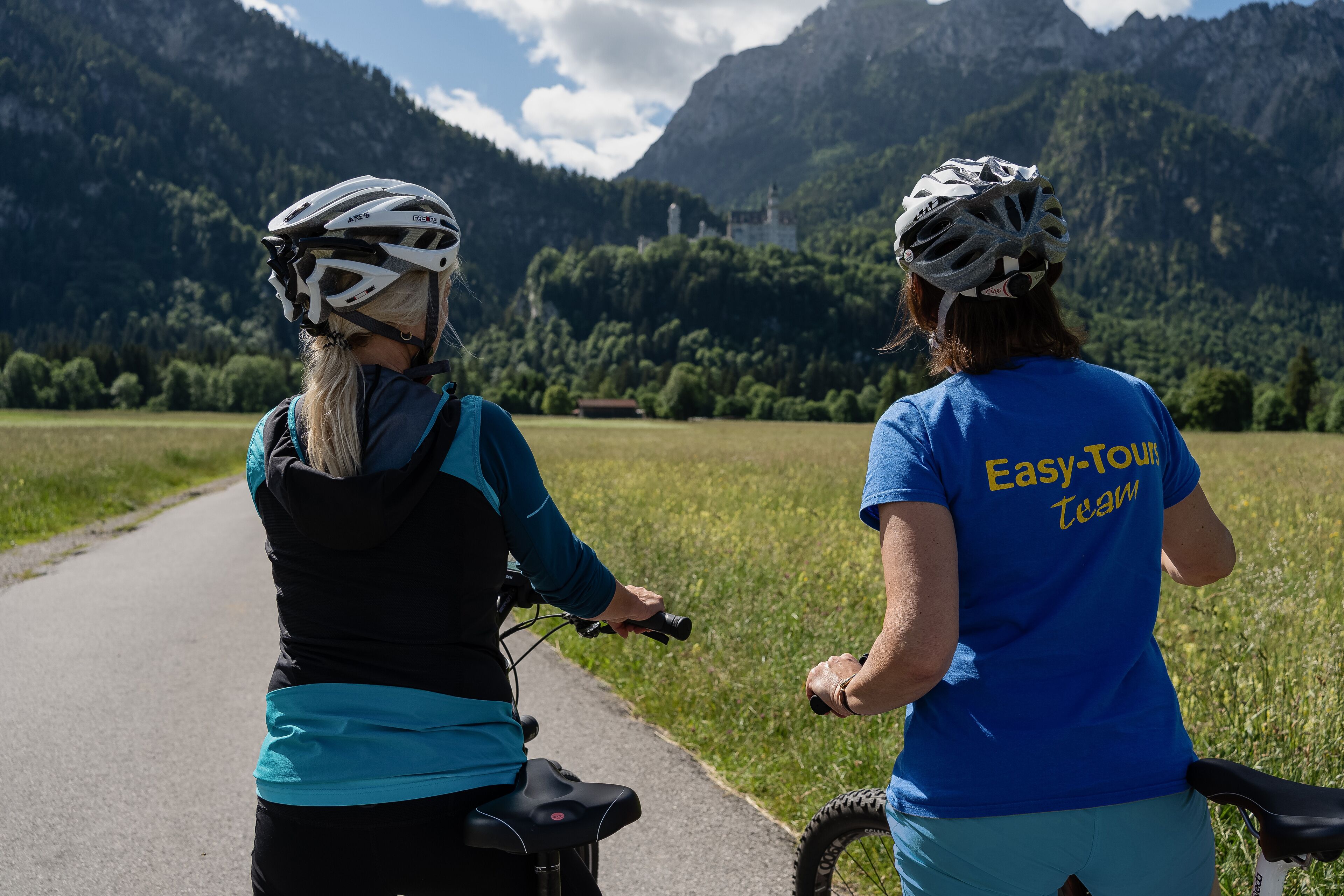 Dois ciclistas em um caminho pavimentado, com uma paisagem montanhosa ao fundo.