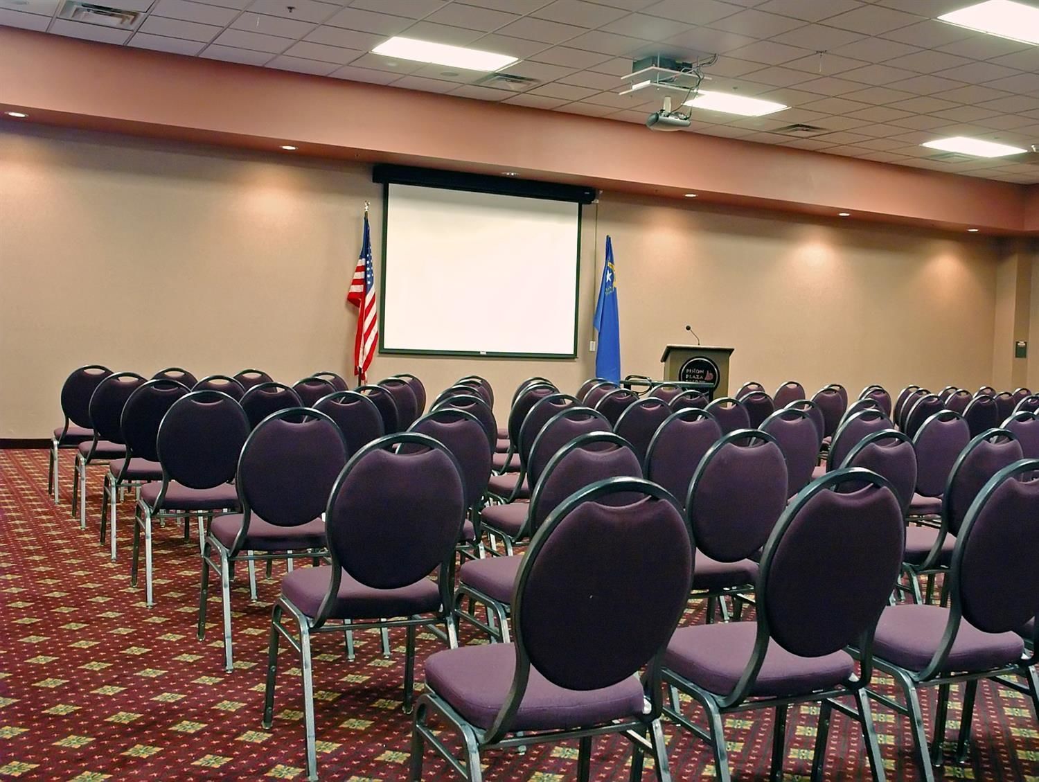 Uma sala de conferências com fileiras de cadeiras roxas, uma tela de projetor, uma bandeira americana e um púlpito com uma placa.