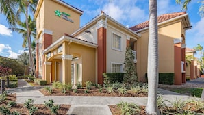 Um hotel com fachada bege, um letreiro com a inscrição "Stay America" e um jardim bem cuidado com palmeiras e arbustos.