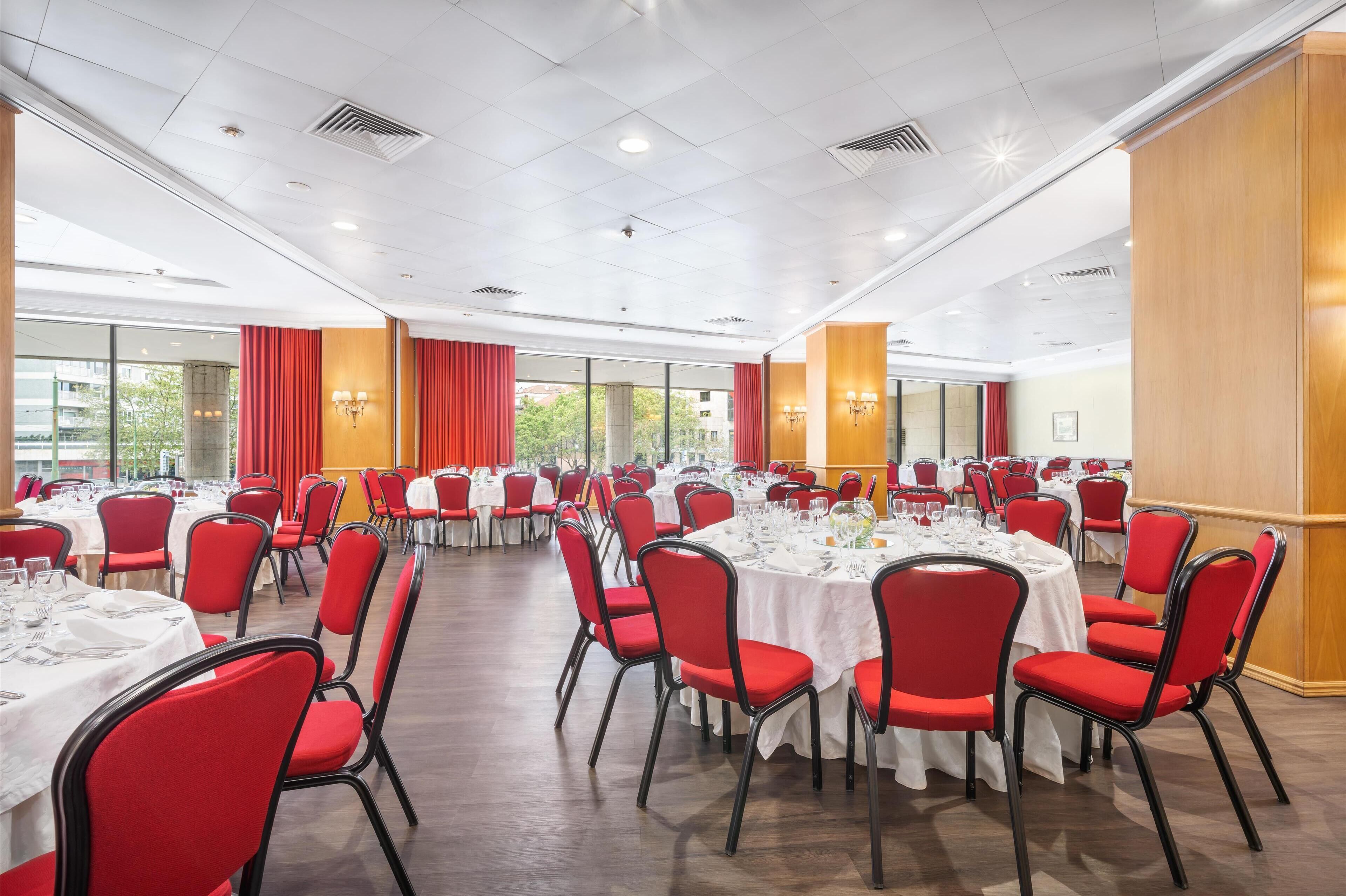 Uma sala de banquetes com mesas redondas, cadeiras vermelhas e toalhas brancas.