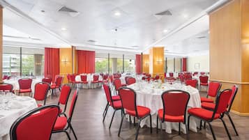 Uma sala de banquetes com mesas redondas, cadeiras vermelhas e toalhas brancas.