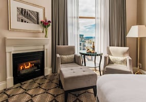 Quarto de hotel com lareira, dois poltronas, uma mesinha de centro com uma garrafa e flores, e vista para a cidade por grandes janelas.