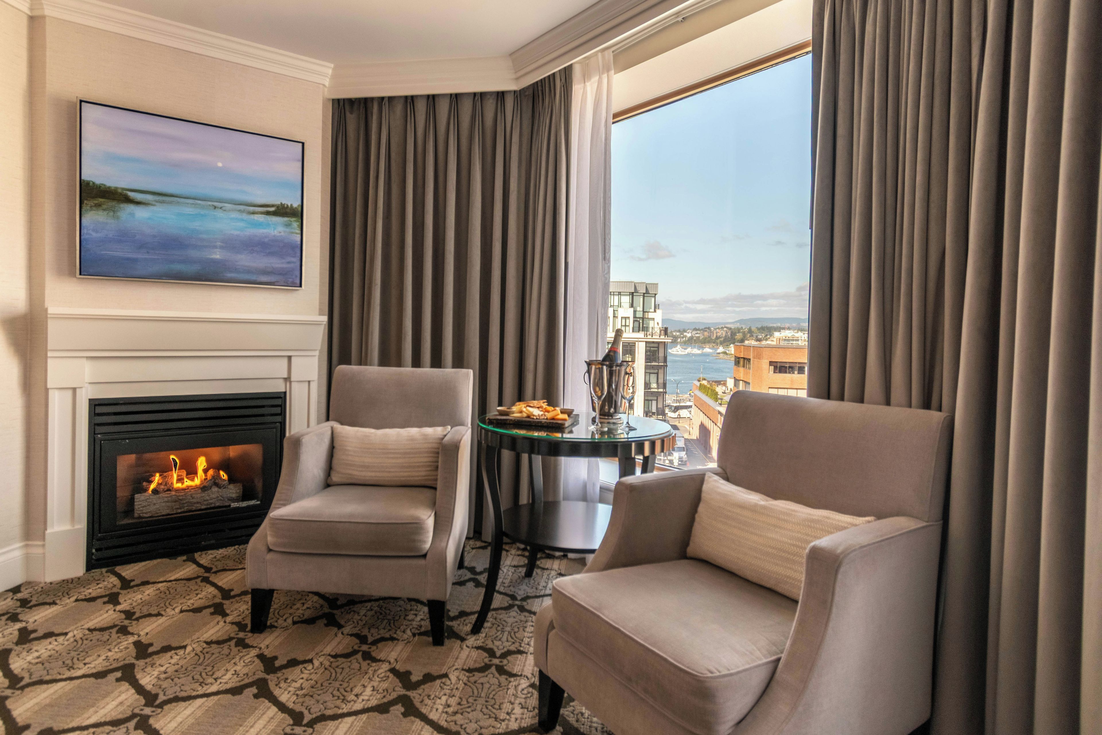 Quarto de hotel com lareira, duas poltronas, uma mesinha com uma garrafa e copos, e um quadro na parede.
