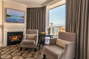 Quarto de hotel com lareira, duas poltronas, uma mesinha com uma garrafa e copos, e um quadro na parede.