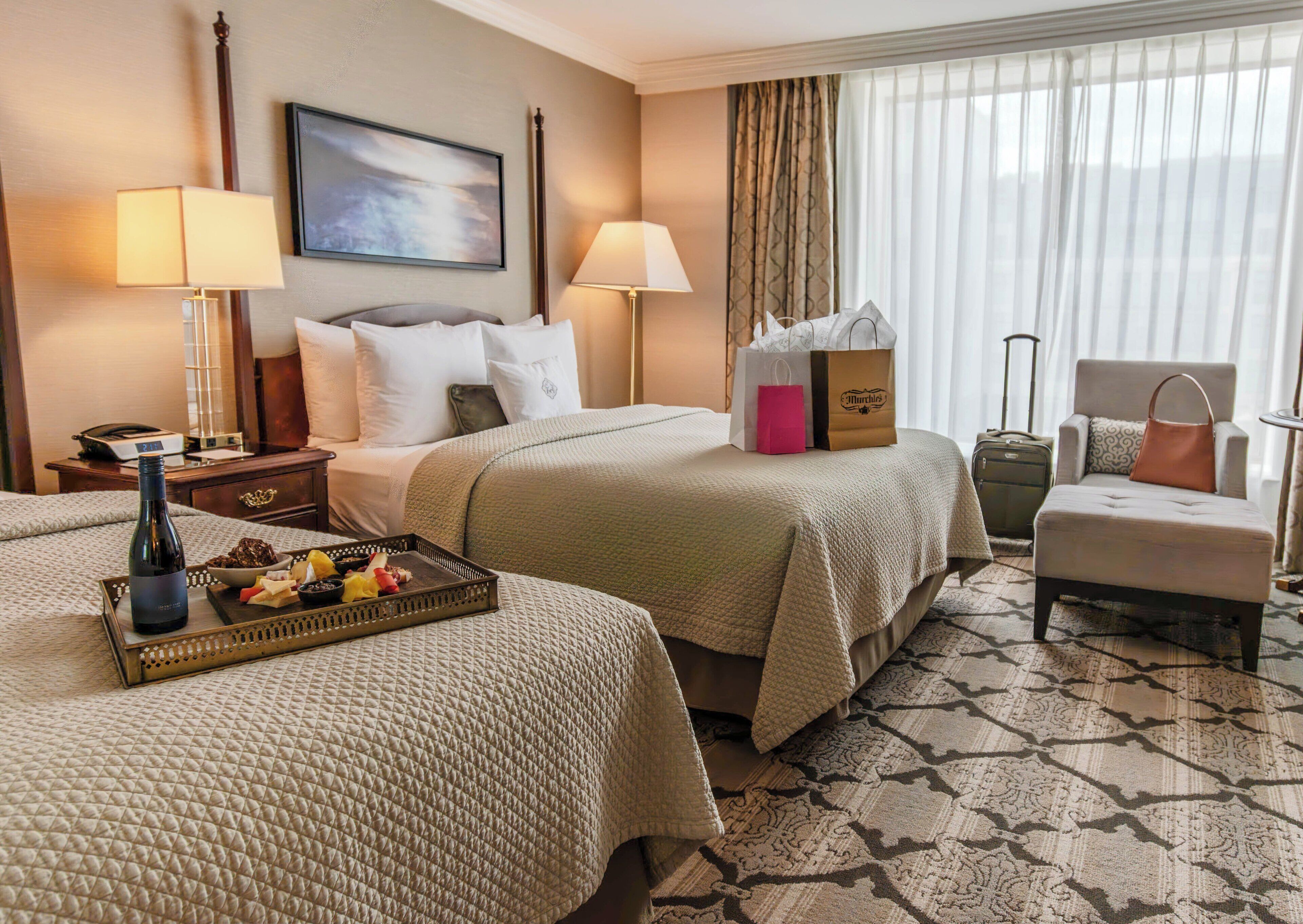 Quarto de hotel com duas camas, uma escrivaninha, uma cadeira, uma mala e um suporte com comida e uma garrafa.