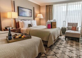 Quarto de hotel com duas camas, uma escrivaninha, uma cadeira, uma mala e um suporte com comida e uma garrafa.