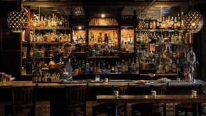 Um bar com um bartender preparando bebidas, prateleiras repletas de garrafas diversas e um interior bem iluminado.
