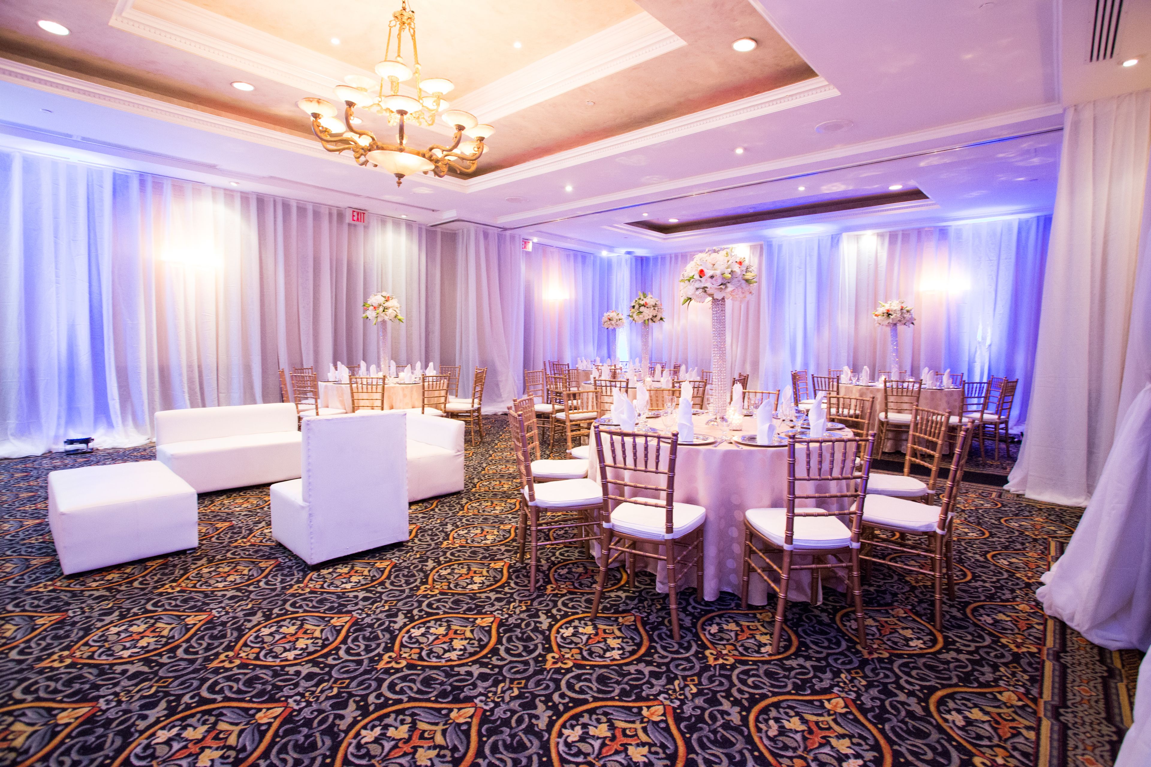 Uma sala de banquetes com mesas redondas, cadeiras brancas e um tapete estampado.