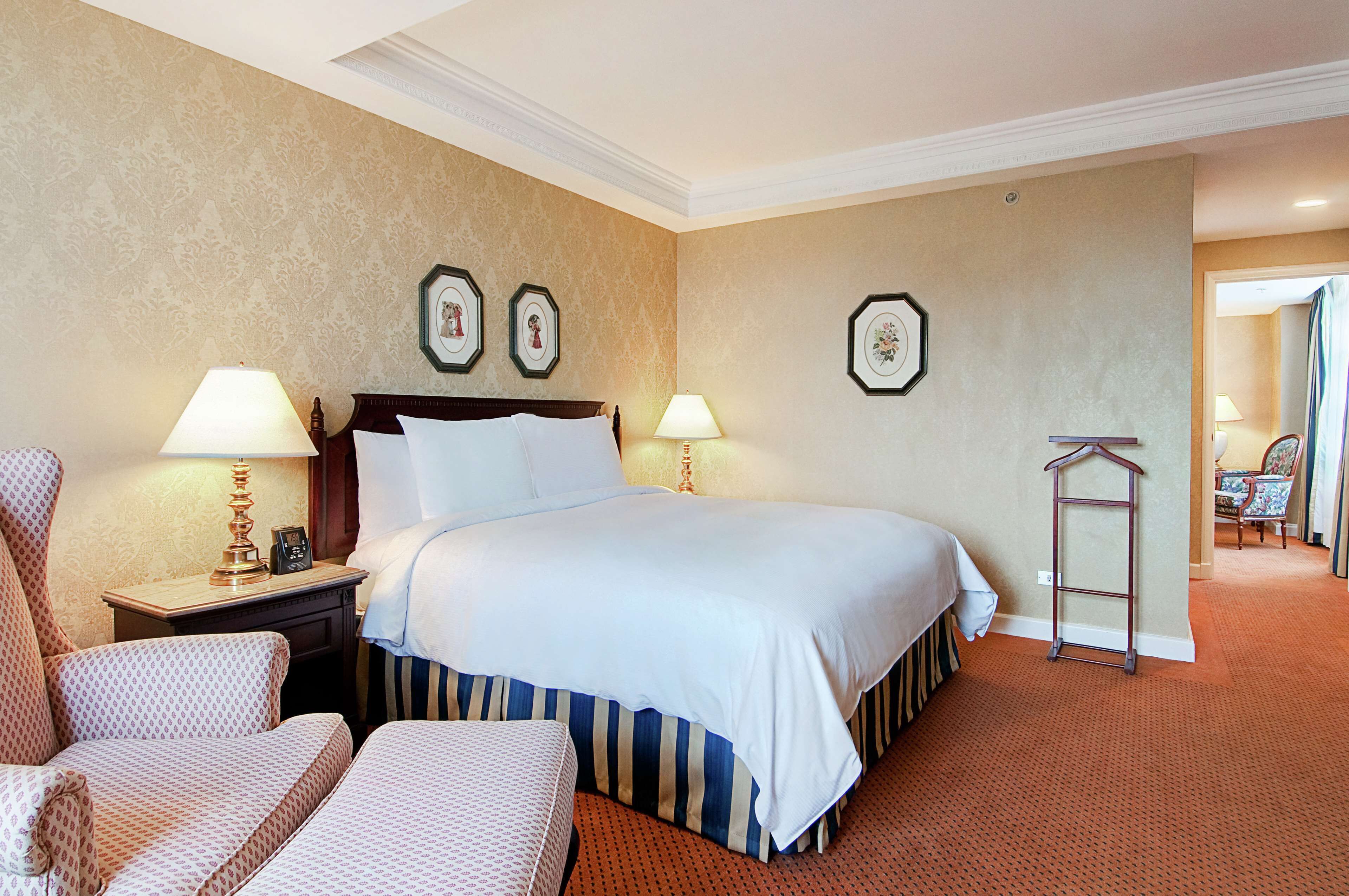 Quarto de hotel com uma cama grande, duas mesas de cabeceira com abajures, uma cadeira e um porta-roupas.