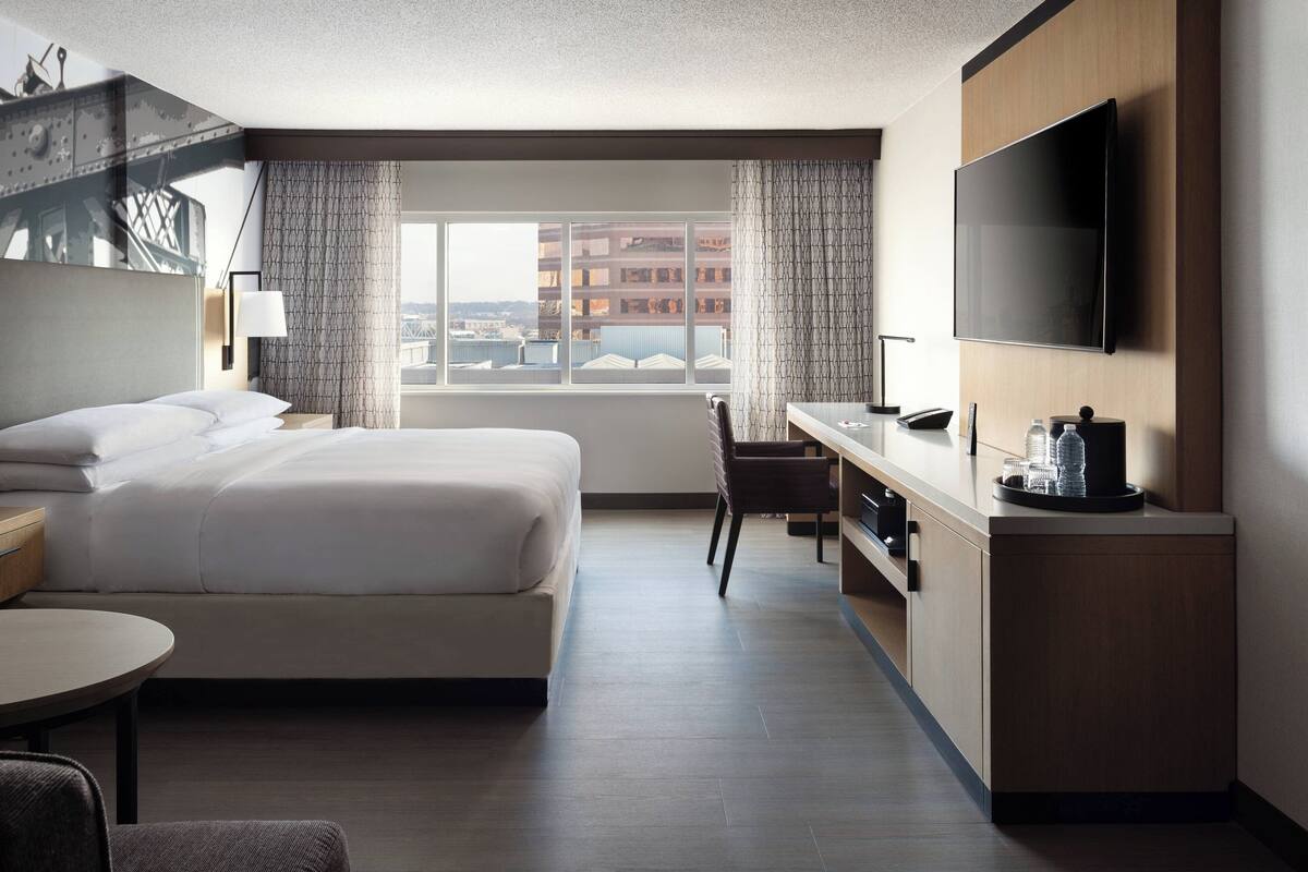Quarto de hotel com uma cama grande, uma escrivaninha, uma cadeira, televisão e janela com vista para a cidade.
