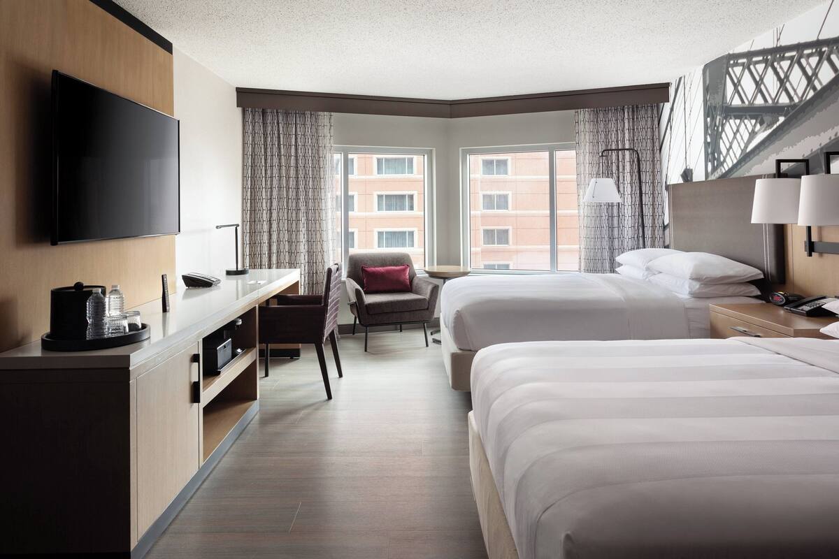 Quarto de hotel com duas camas, uma escrivaninha com televisão de tela plana, uma cadeira e vista para edifícios através de amplas janelas.