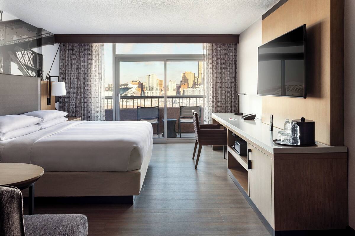 Quarto de hotel com uma cama grande, uma escrivaninha, uma televisão e varanda com vista para a cidade.