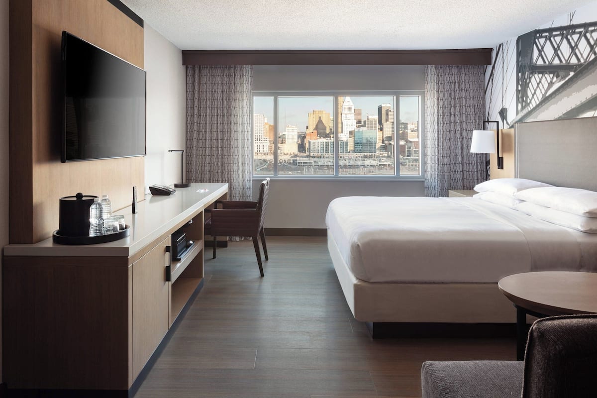 Quarto de hotel com uma cama grande, escrivaninha com televisão de tela plana, vista para a cidade pela janela e design moderno.