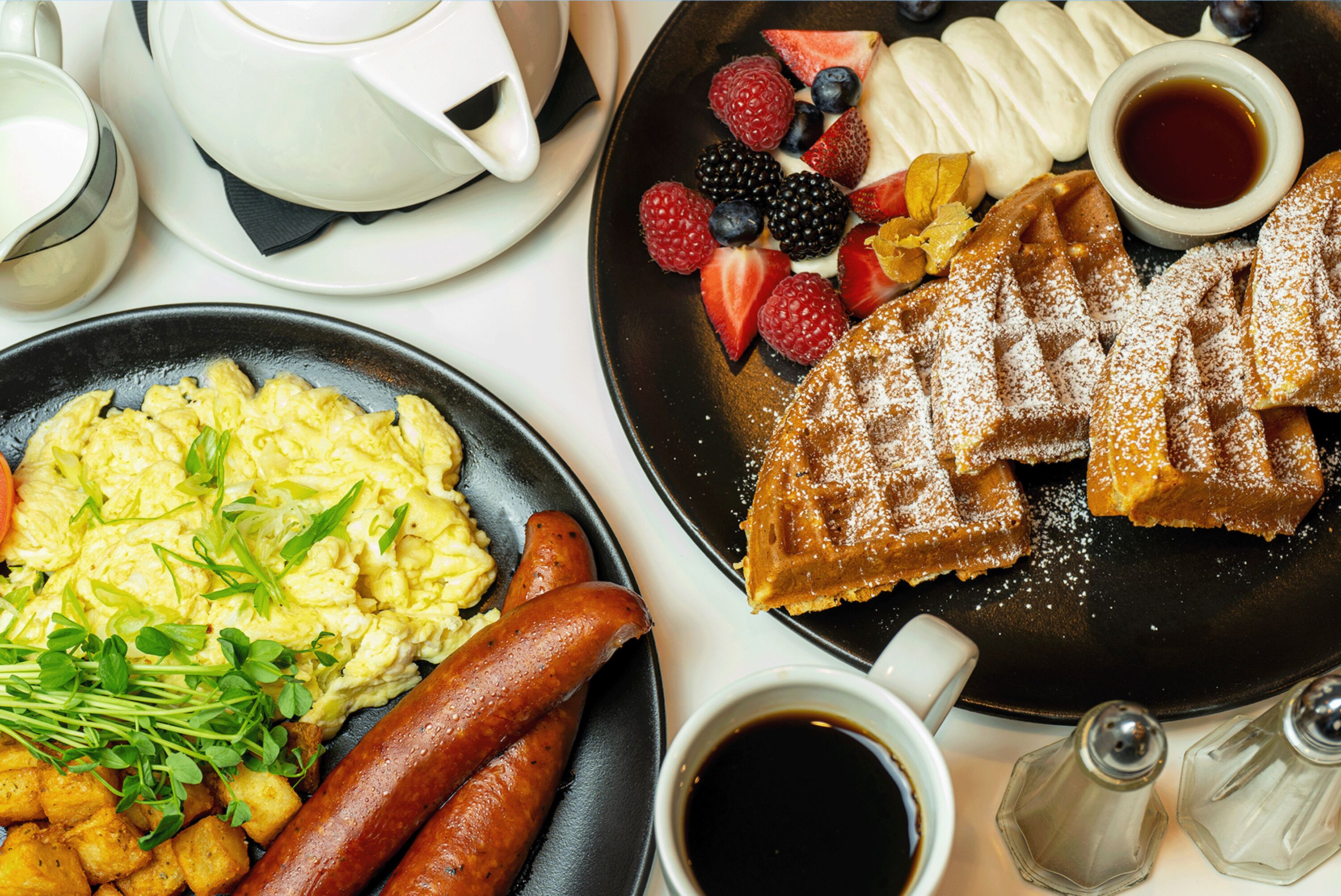 Prato de café da manhã com ovos mexidos, salsichas e hash browns, um waffle com frutas e xarope, e uma xícara de café.