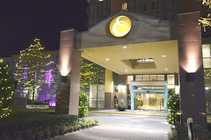 Entrada de hotel com um logotipo iluminado e chamativo, porta de vidro e placa indicando que o hotel está aberto.