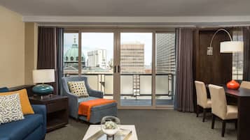 Quarto de hotel com sofá, poltrona, mesa de jantar e vista para a cidade através de grandes janelas.
