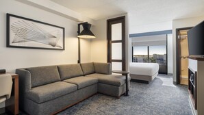 Quarto de hotel moderno com sofá seccional cinza, mesa lateral pequena, luminária de chão, janela grande com vista para a cidade e cama com televisão.