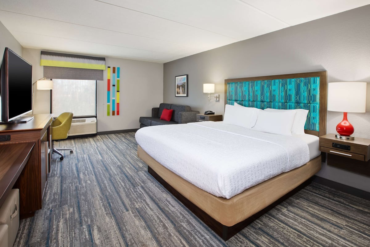 Quarto de hotel com uma cama grande, uma escrivaninha com cadeira, televisão e um sofá com almofada vermelha.