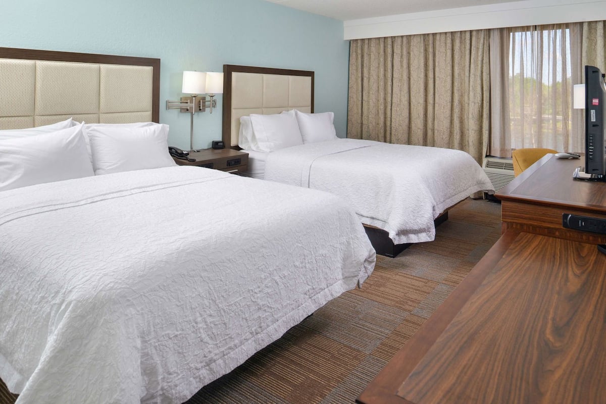 Quarto de hotel com duas camas, uma escrivaninha, uma luminária, uma televisão e uma janela com cortinas.