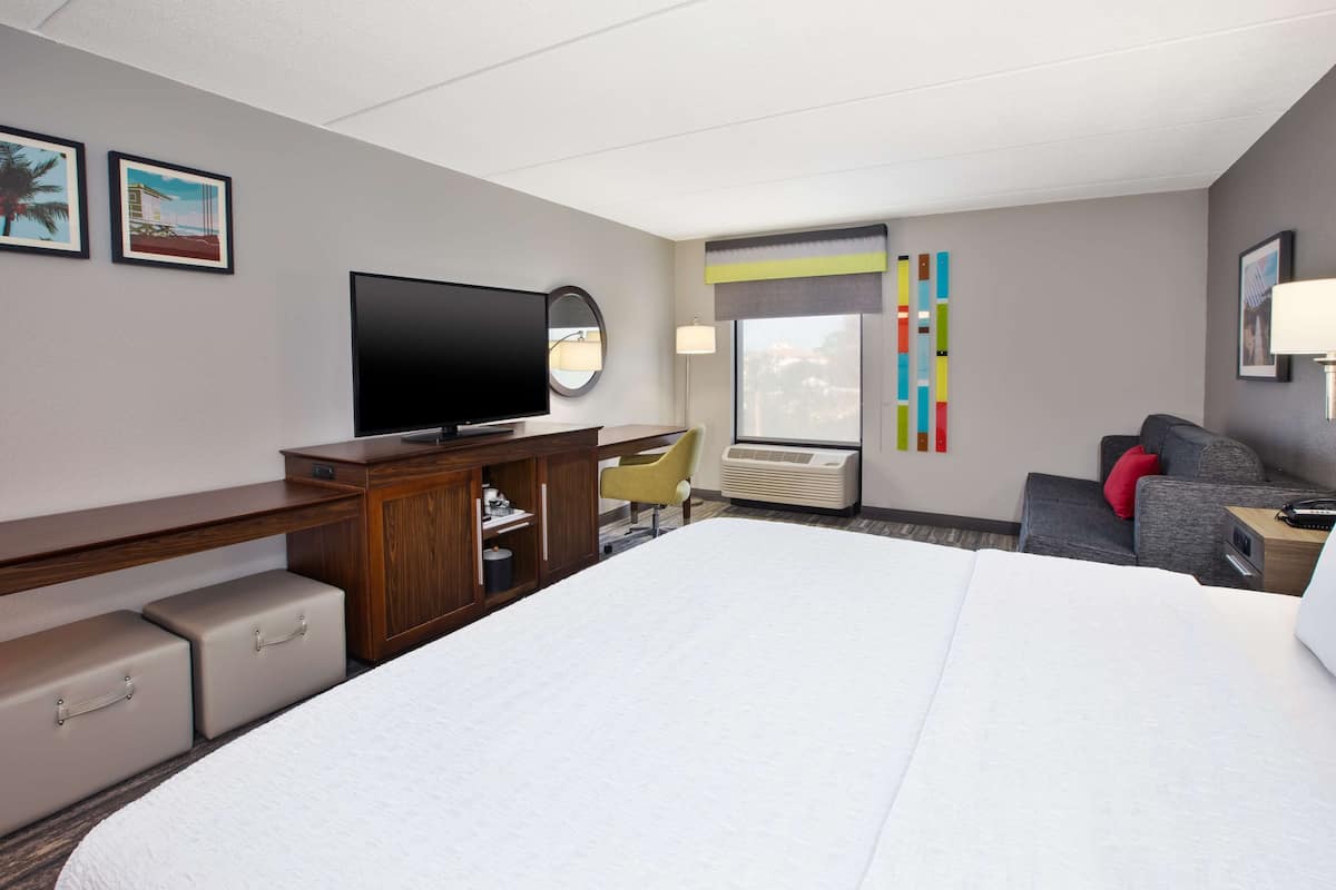 Quarto de hotel com uma cama grande, uma escrivaninha com televisão de tela plana, uma poltrona, um sofá e uma janela com vista.