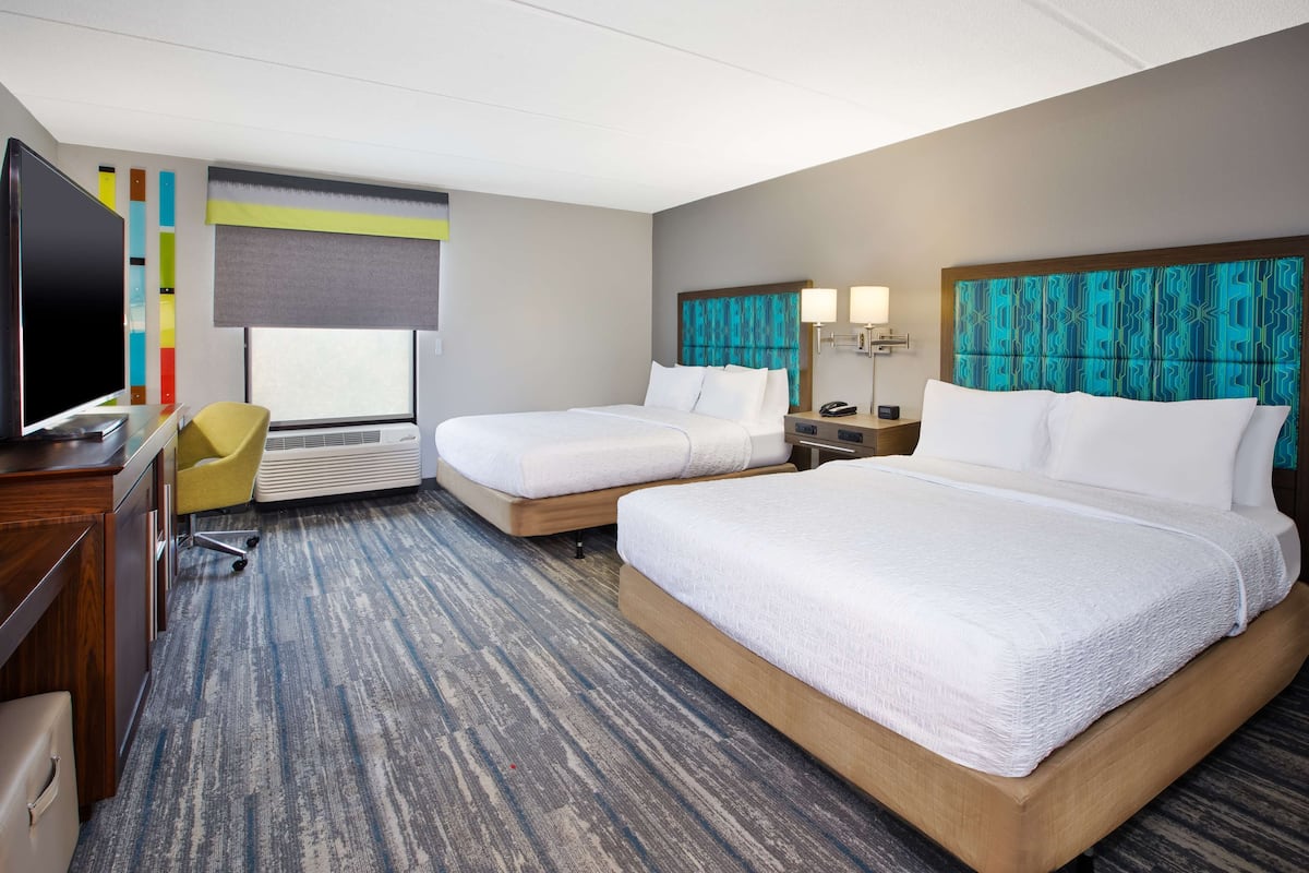 Quarto de hotel com duas camas, uma escrivaninha com televisão, uma cadeira e janela com vista.
