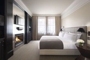 Quarto de hotel com lareira, cama com roupa de cama branca, mesa de cabeceira com abajur e vista da cidade por grandes janelas.