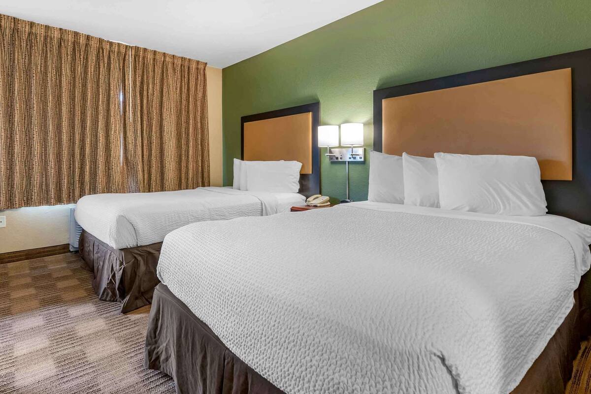 Quarto de hotel com duas camas, uma janela grande com cortinas e uma televisão de tela plana na parede.