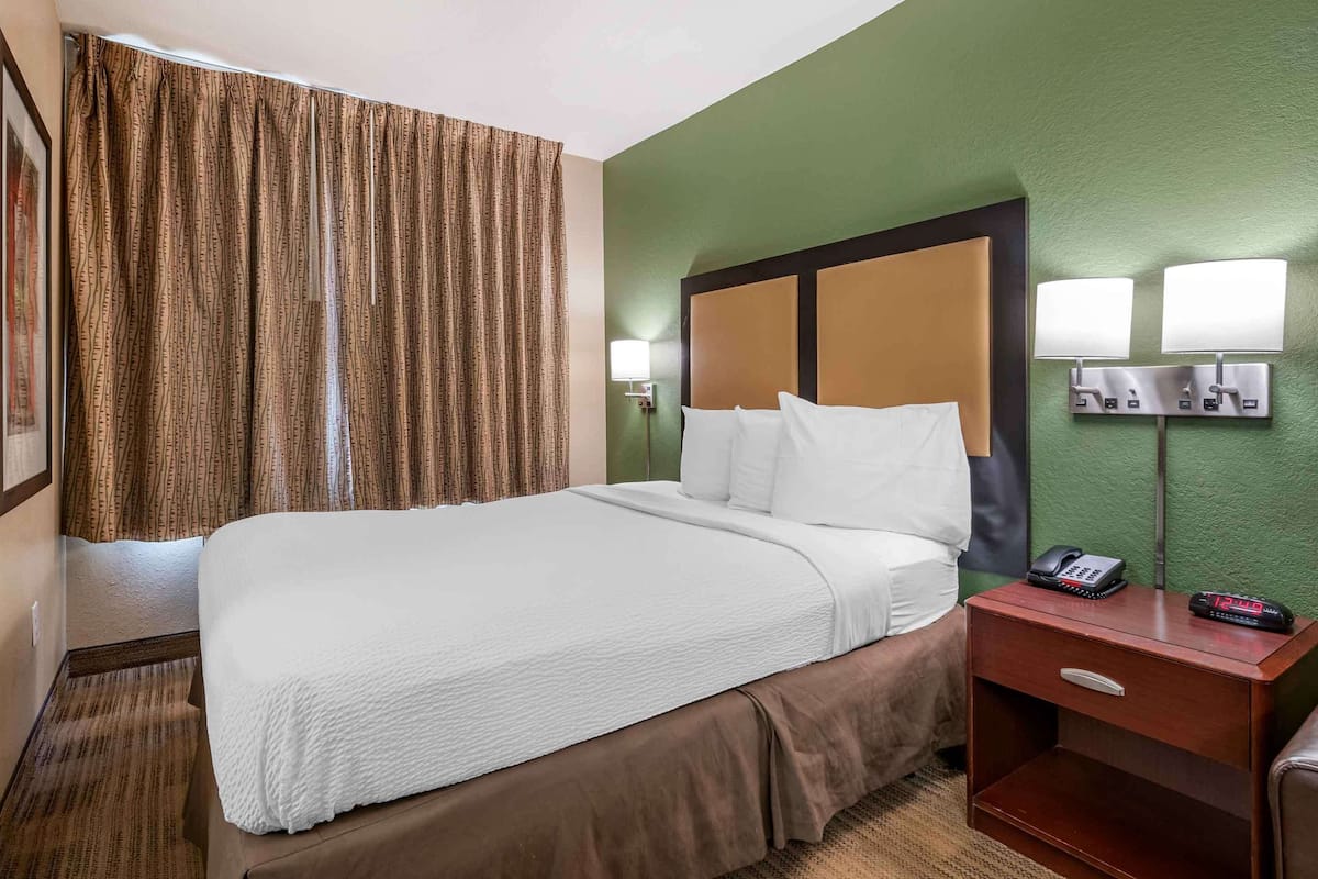 Quarto de hotel com uma cama grande, uma mesa de cabeceira com telefone e relógio, uma parede verde e uma janela com cortinas.