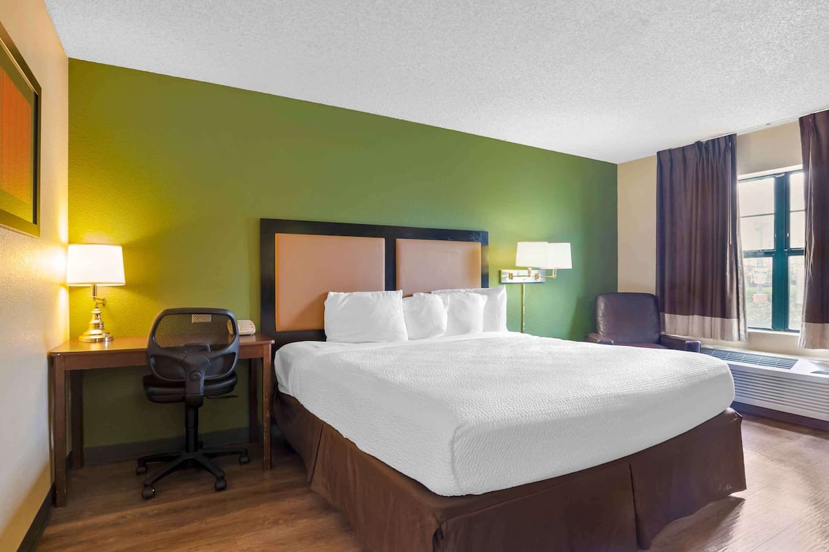 Quarto de hotel com uma cama grande, uma escrivaninha com cadeira, duas luminárias de cabeceira e uma janela com cortinas.