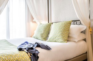 Cama bem arrumada com almofadas estampadas, um dossel translúcido e uma peça de roupa dobrada sobre a cama.