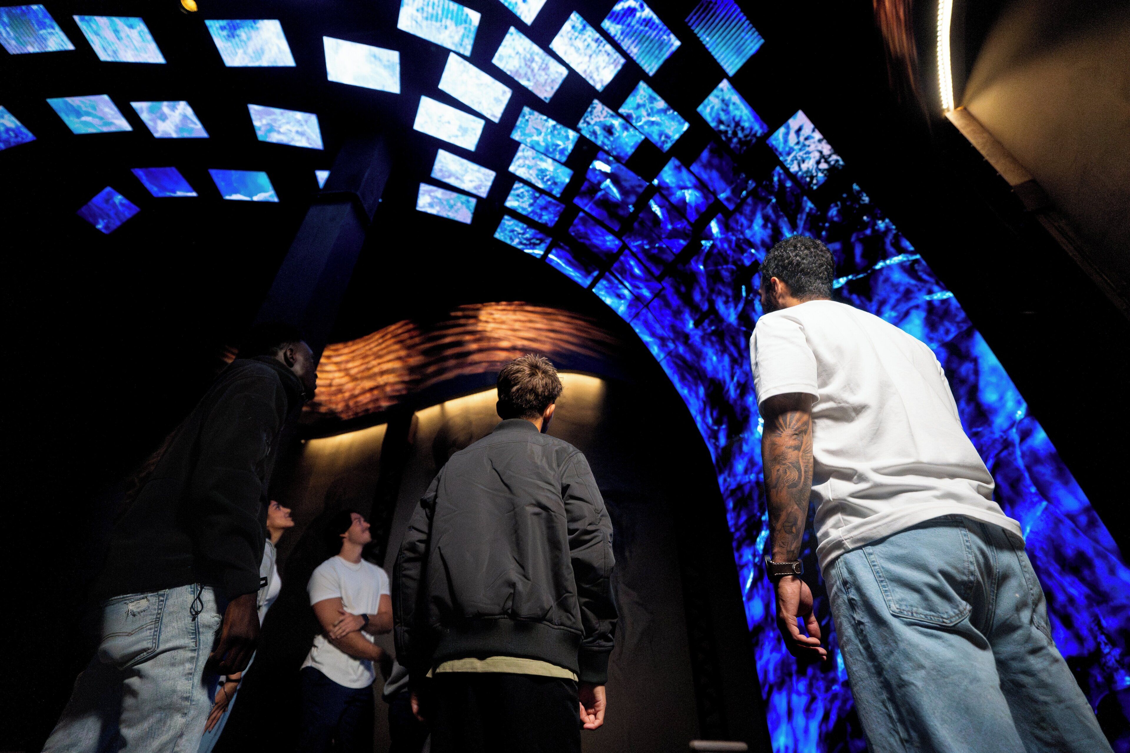 Um grupo de pessoas caminhando por um túnel com um painel digital no teto.
