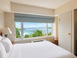 Quarto de hotel com uma janela grande, uma cama, uma mesinha de cabeceira, um abajur e uma porta.